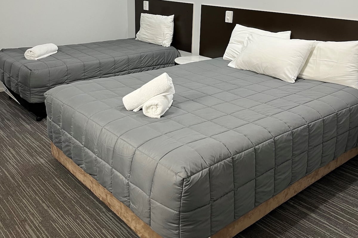 Quarto de hotel com duas camas, cada uma com colcha cinza acolchoada e travesseiros brancos, um ventilador de teto e um quadro na parede.
