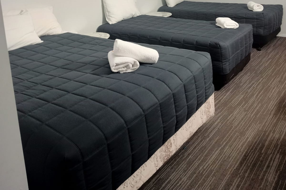Quarto de hotel com três camas escuras, almofadas brancas e um ventilador de teto.