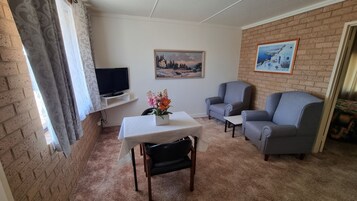 Uma sala de estar com televisão, dois poltronas, uma mesa com um vaso de flores e um quadro na parede.