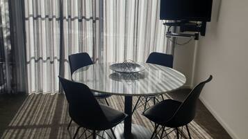 Quarto com uma mesa de vidro redonda, quatro cadeiras pretas, televisão de tela plana na parede e cortinas leves.