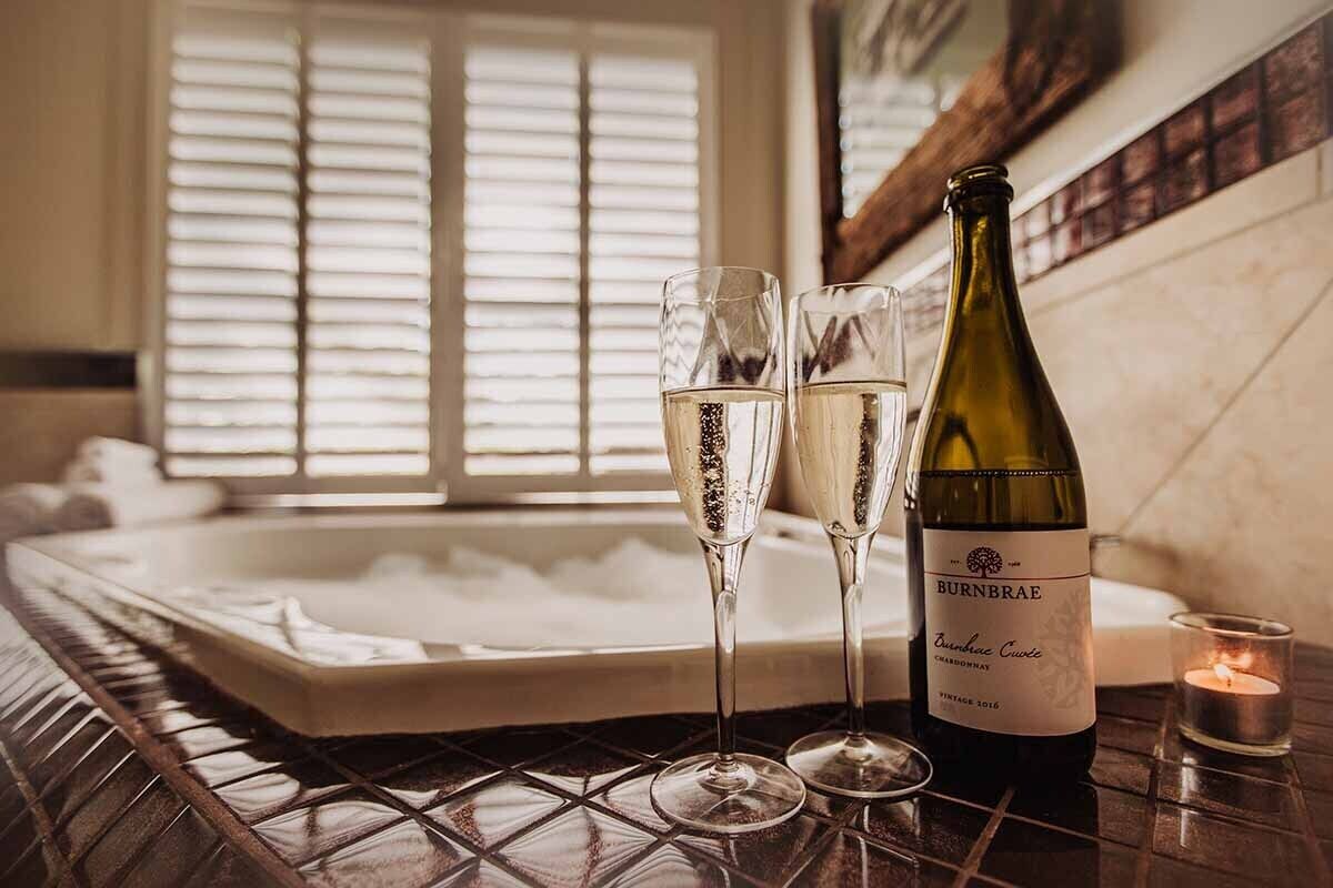 Uma garrafa de Burnbrae Chardonnay, duas taças de vinho e uma vela sobre uma mesa em um quarto com banheira e janela com persianas.