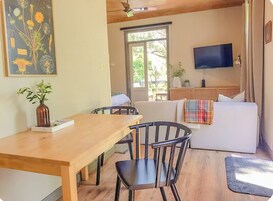 Uma sala de estar acolhedora com uma mesa de jantar de madeira, cadeiras pretas, um sofá branco e uma televisão fixada na parede.