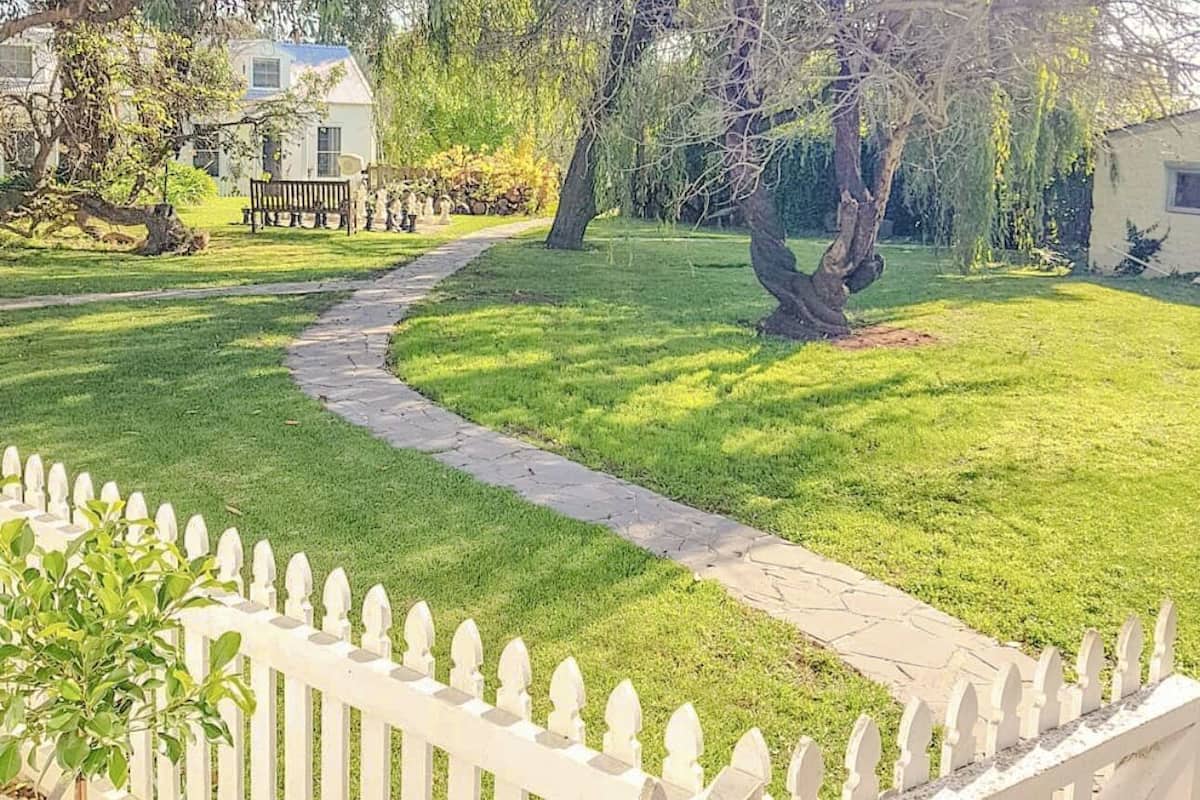 Um gramado bem cuidado com um caminho sinuoso, uma cerca branca de piquete e um banco ao fundo.