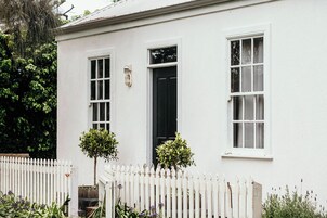 Um prédio branco com duas janelas e uma porta, cerca branca de piquete e pequenas árvores na frente.
