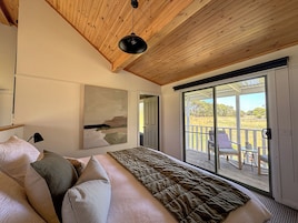Quarto com cama, quadro, escrivaninha e vista para um deck externo por uma porta deslizante.