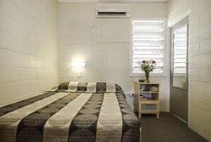 Uma cama de solteiro com colcha estampada, luminária de cabeceira, aparelho de ar condicionado, janela com persianas, pequena estante de madeira com um vaso de flores e uma porta.