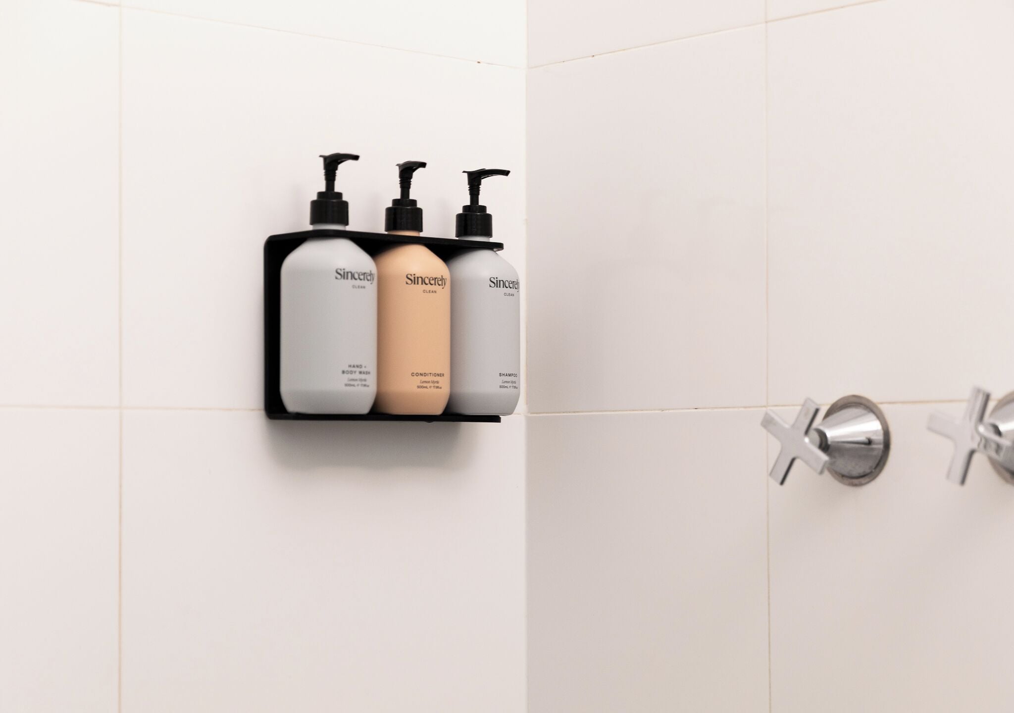 Um banheiro com uma prateleira contendo três frascos de produtos Sincerely para o corpo, condicionador e shampoo.