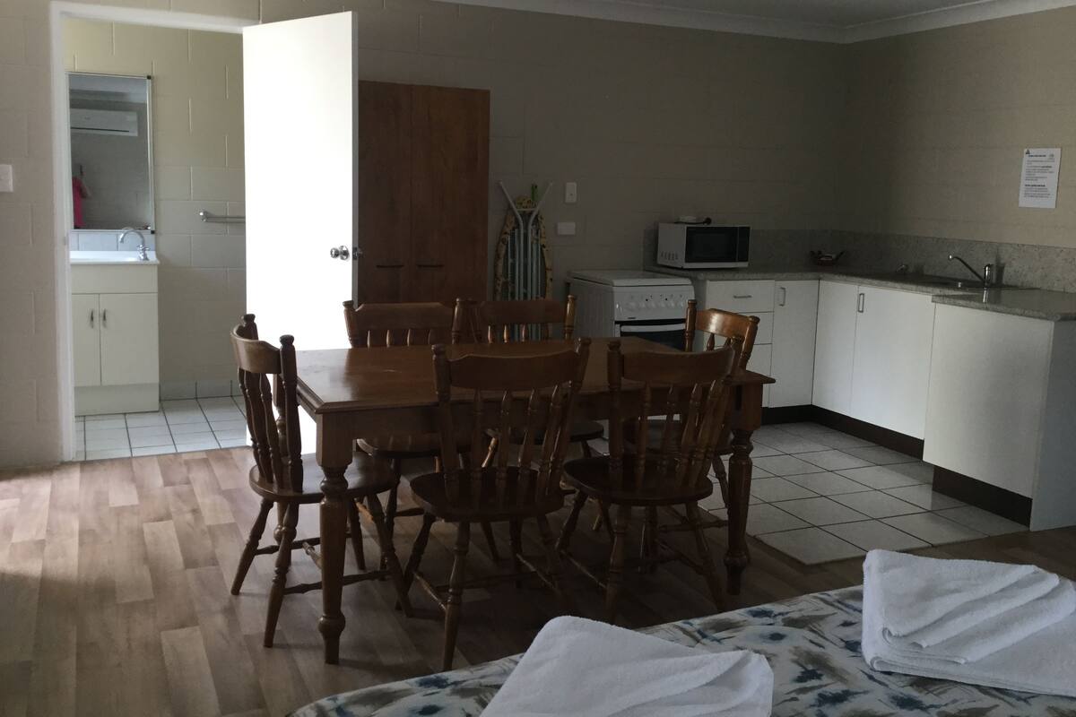 Uma área de refeições com mesa e cadeiras de madeira, uma cozinha com armários e eletrodomésticos brancos, e uma porta visível que dá para outro cômodo.