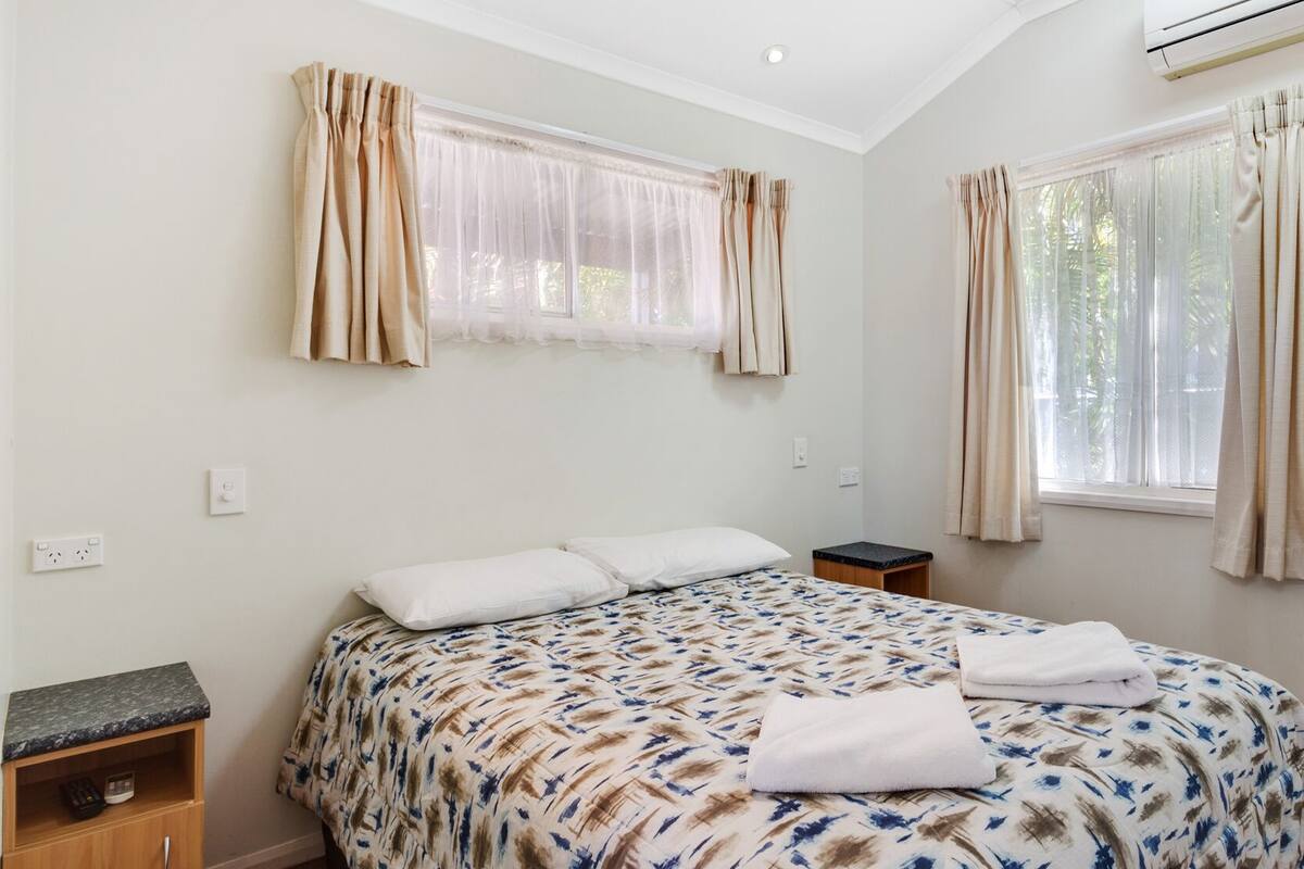 Quarto com cama, duas mesas de cabeceira, um criado-mudo, janela com cortinas e aparelho de ar condicionado.
