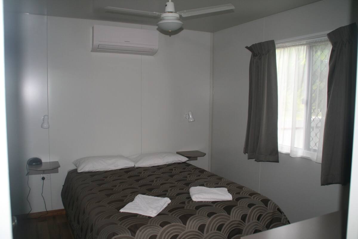Quarto com cama, ventoinha no teto, ar condicionado, mesa de cabeceira com abajur e janela com cortinas.