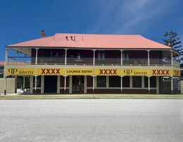 Um edifício de dois andares com telhado vermelho e varandas. Apresenta placas com 'QP Tavern' e 'XXXX Lounge Entrada'.