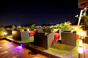 Espaço ao ar livre com mobiliário de vime e toalhas de mesa vermelhas, rodeado por plantas em vasos e iluminado por iluminação ambiente.