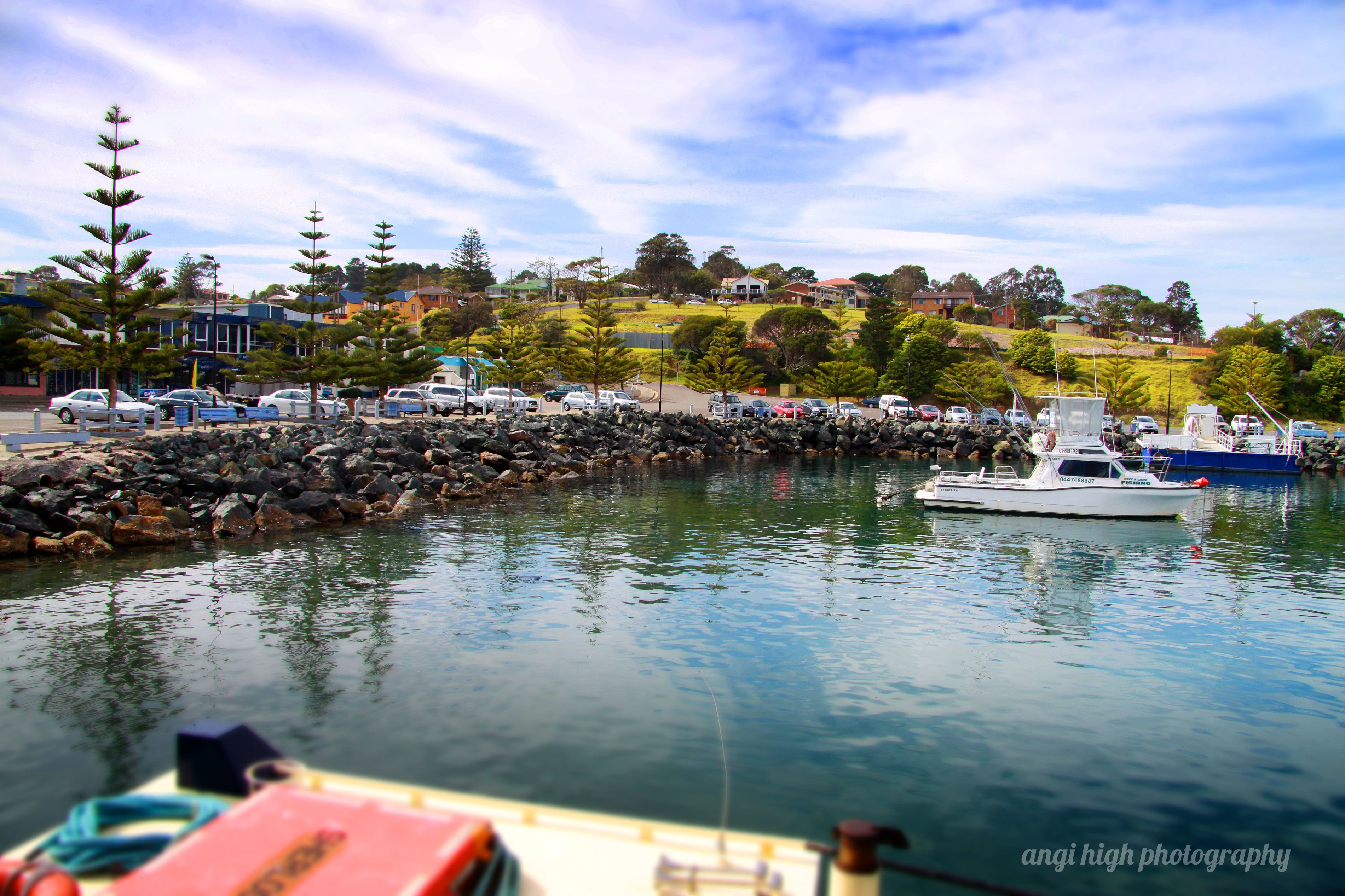 Uma marina com barcos atracados, um molhe rochoso e casas ao fundo.