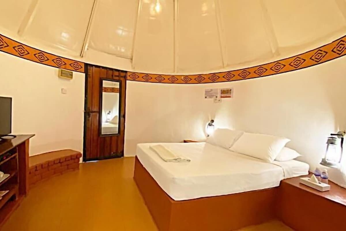 Quarto com cama, mesa de cabeceira, abajur e televisão sobre um suporte de madeira.