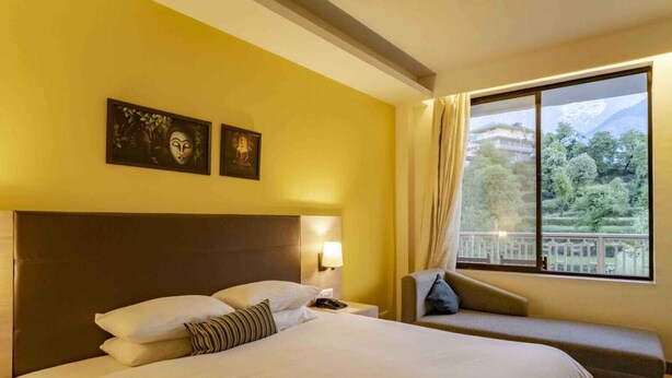 Quarto de hotel com uma cama grande, um sofá, uma mesa de cabeceira com abajur e uma janela com vista para áreas verdes e um edifício.