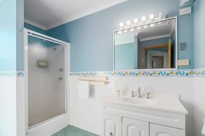 Casa de banho com chuveiro, pia com duas torneiras, espelho com iluminação e painel de azulejos.