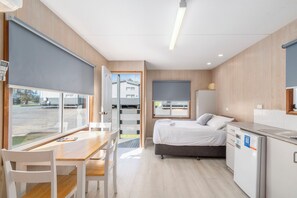 Quarto de hotel compacto com cama, pequena cozinha, área de jantar com mesa e cadeiras, e uma janela grande com vista para o exterior.
