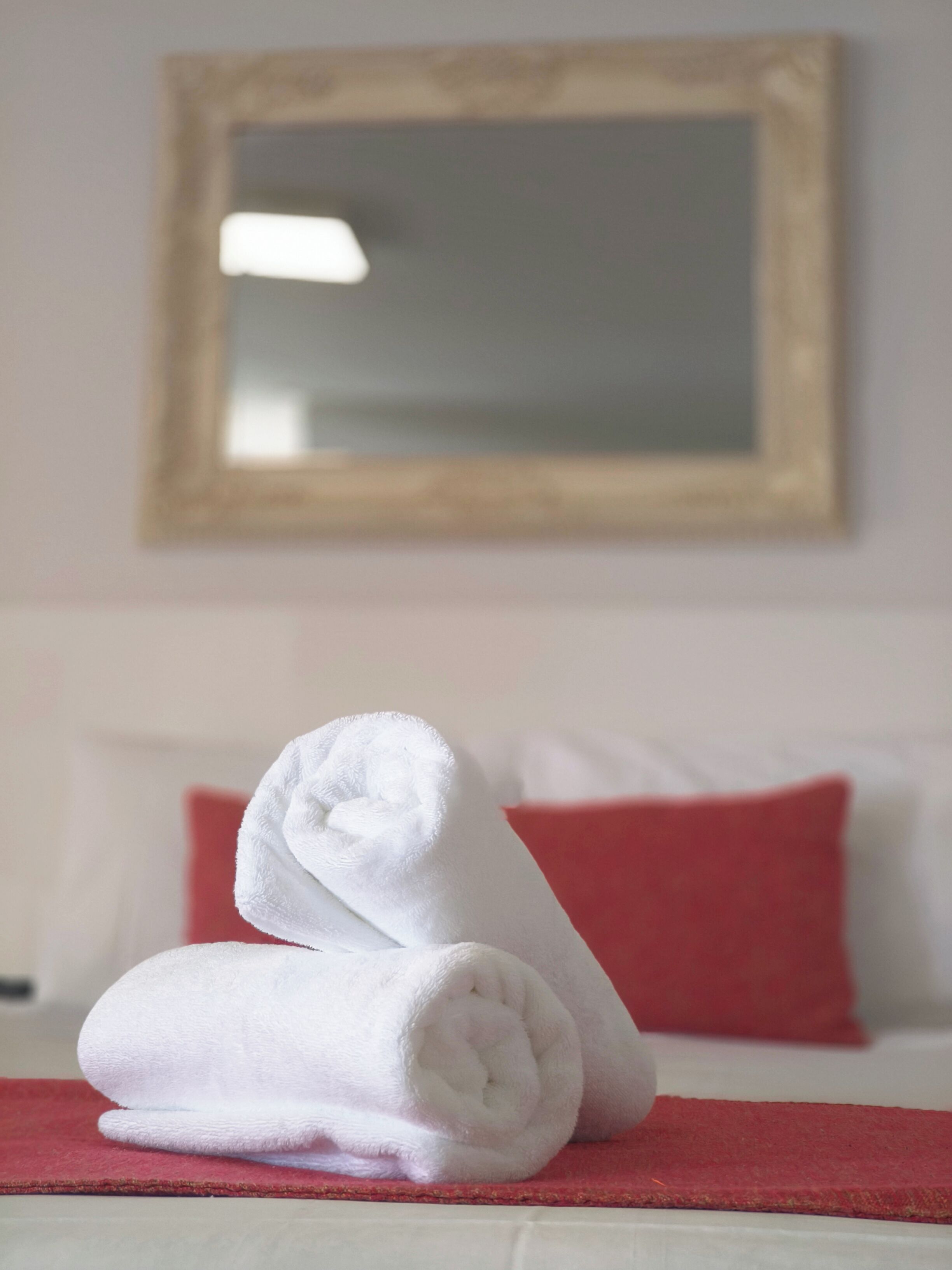 Cama bem arrumada com roupa de cama branca e vermelha, uma toalha branca dobrada e um espelho na parede.