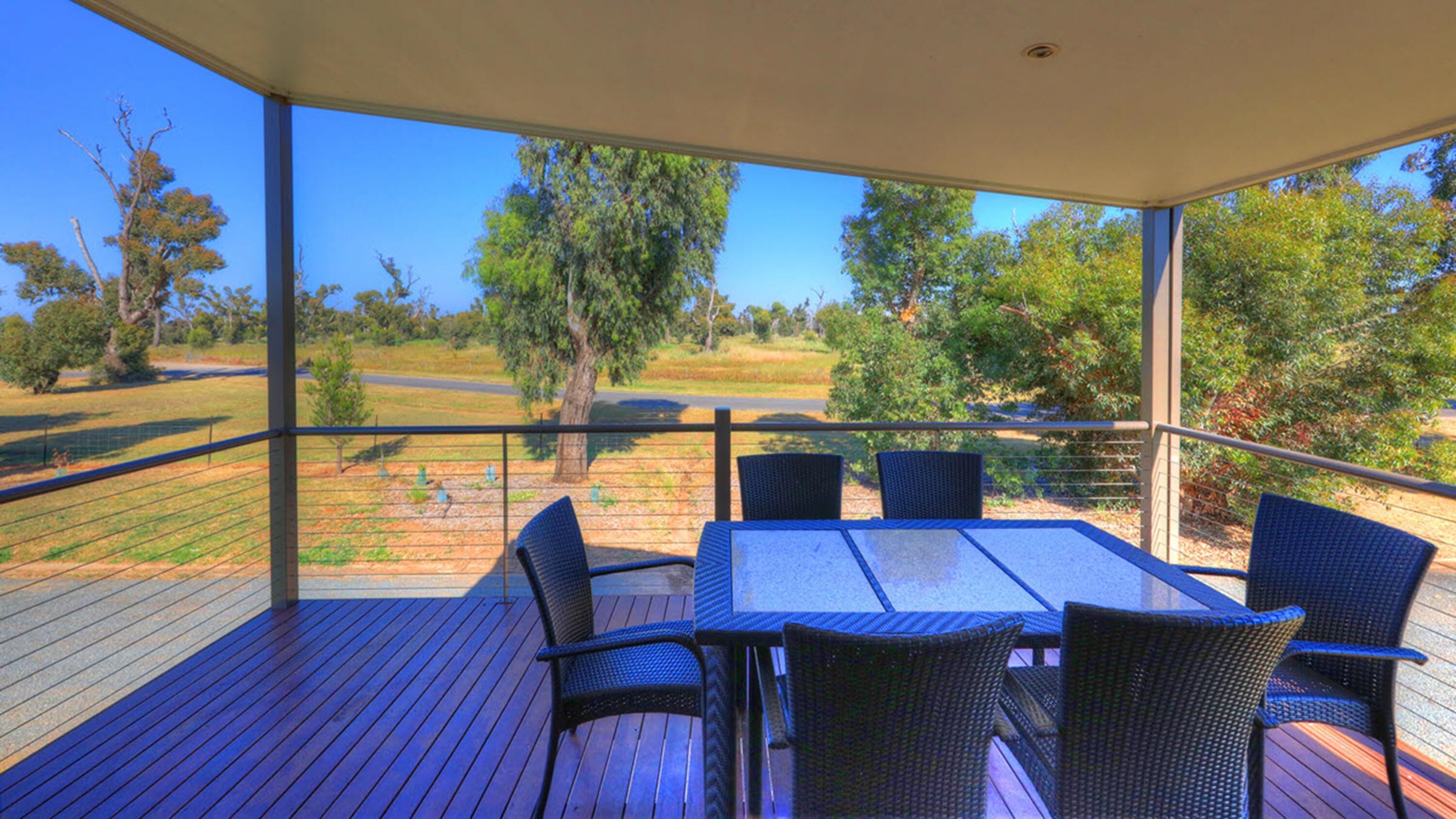 Uma área externa coberta com mesa e cadeiras, com vista para um campo gramado e árvores.