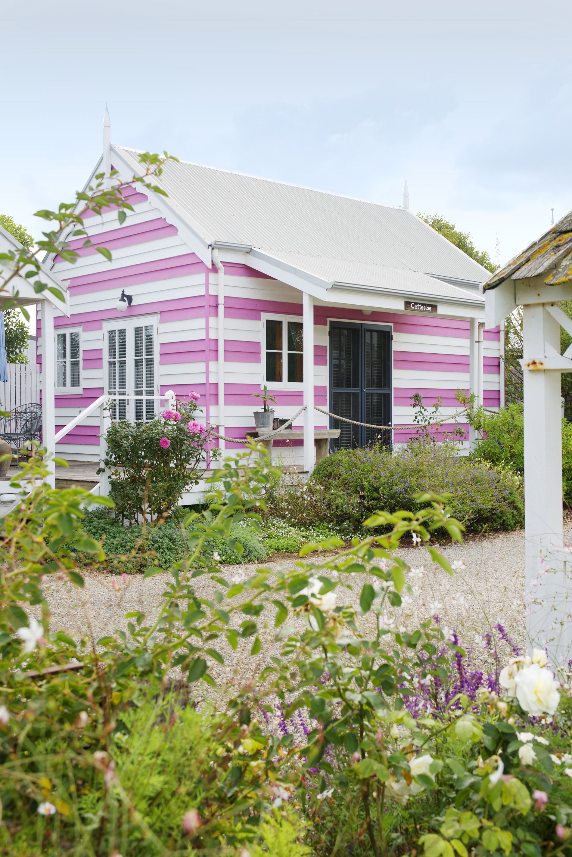 Uma casa listrada em rosa e branco, com porta preta, cercada por um jardim com diversas plantas e flores.