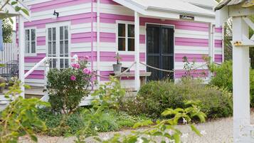 Uma casa listrada em rosa e branco, com porta preta, cercada por um jardim com diversas plantas e flores.