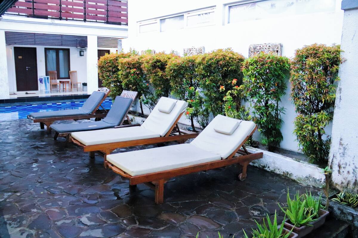 Uma área à beira da piscina com espreguiçadeiras, piso de pedra e um edifício branco com janelas e porta.