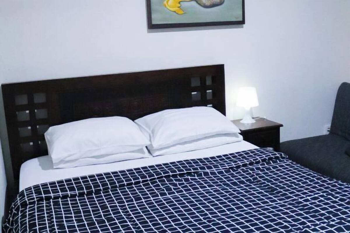 Uma cama com cabeceira escura, almofadas brancas e colcha estampada.