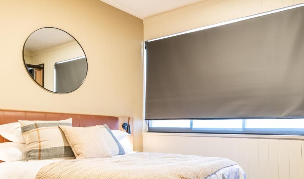 Quarto de hotel com cama, espelho redondo, janela com persiana e radiador.