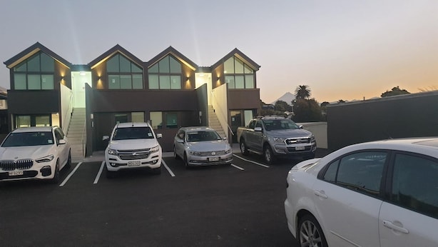 Um estacionamento com cinco carros estacionados em frente a um prédio moderno com janelas grandes e telhado inclinado.