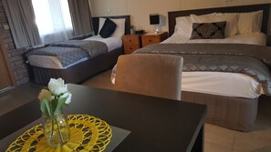 Quarto de hotel com duas camas, uma mesa de jantar e um vaso de flores, e uma parede de tijolos.