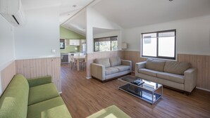 Ampla sala de estar com sofá verde, sofás bege e mesa de centro de vidro. Uma ventoinha de teto e grandes janelas proporcionam iluminação natural.
