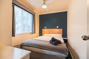 Quarto com cama, mesa de cabeceira, ventilador de teto e janela com cortinas.