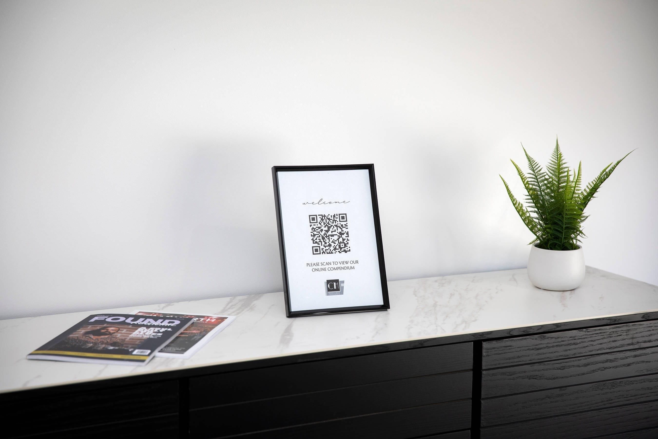 Placa de boas-vindas emoldurada com um código QR num balcão de mármore branco, uma revista intitulada "FOUND" e uma planta em vaso.