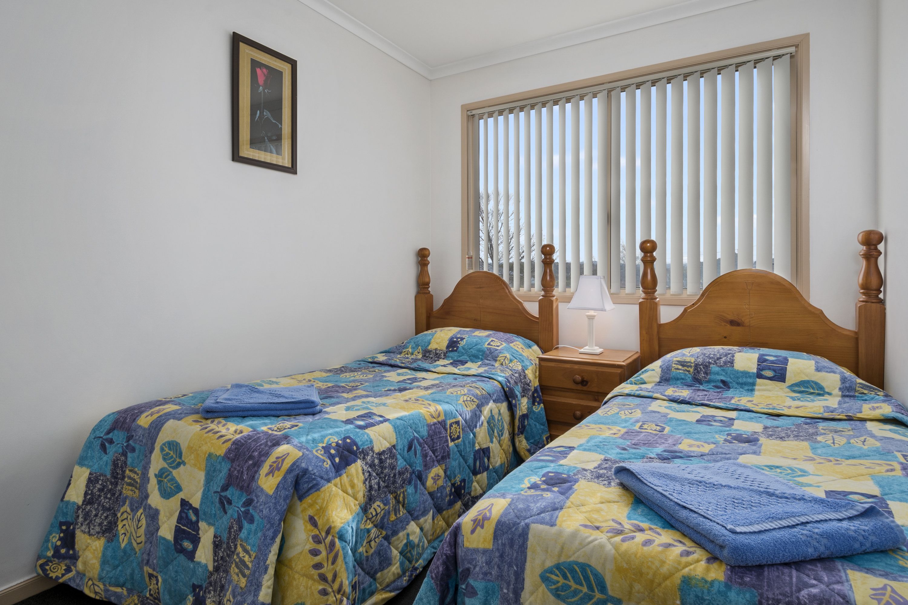 Quarto com duas camas de madeira, uma mesa de cabeceira, um quadro na parede e uma janela com persianas.