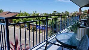 Varanda com vista para um estacionamento e edifícios, com um letreiro azul, plantas em vasos e móveis de exterior.