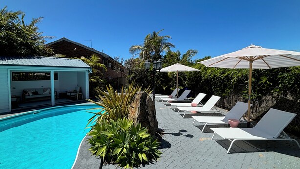 Piscina com espreguiçadeiras e guarda-sóis, cercada por vegetação e área de estar coberta.