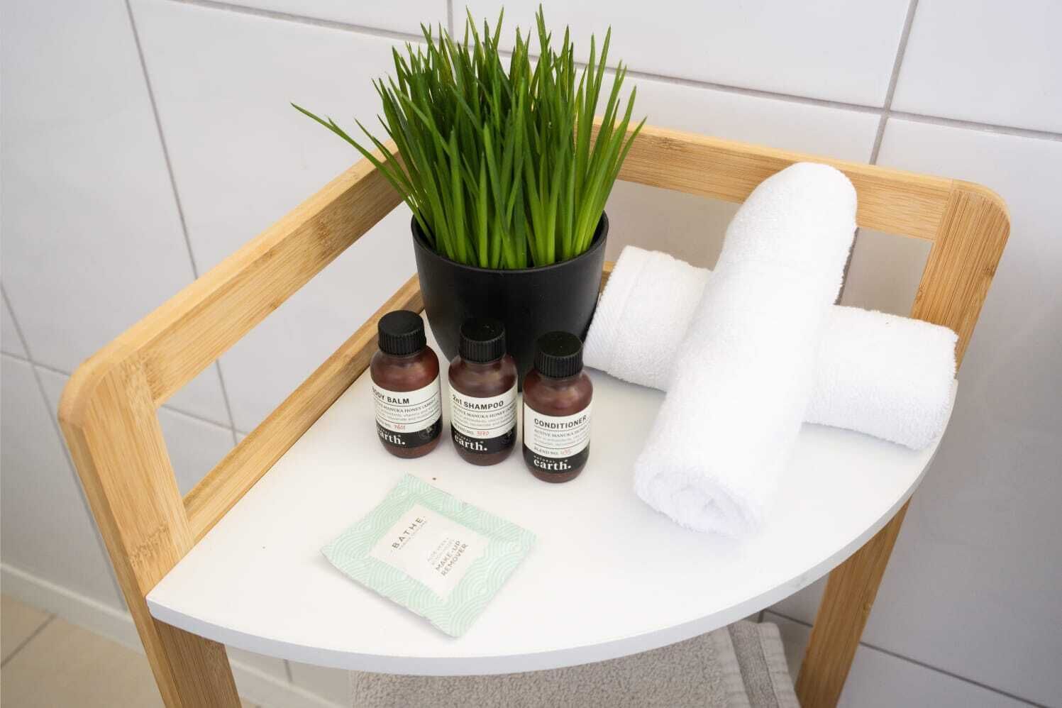 Casa de banho com uma cadeira de madeira, um vaso de planta, uma toalha branca e três frascos de produtos para cabelo.