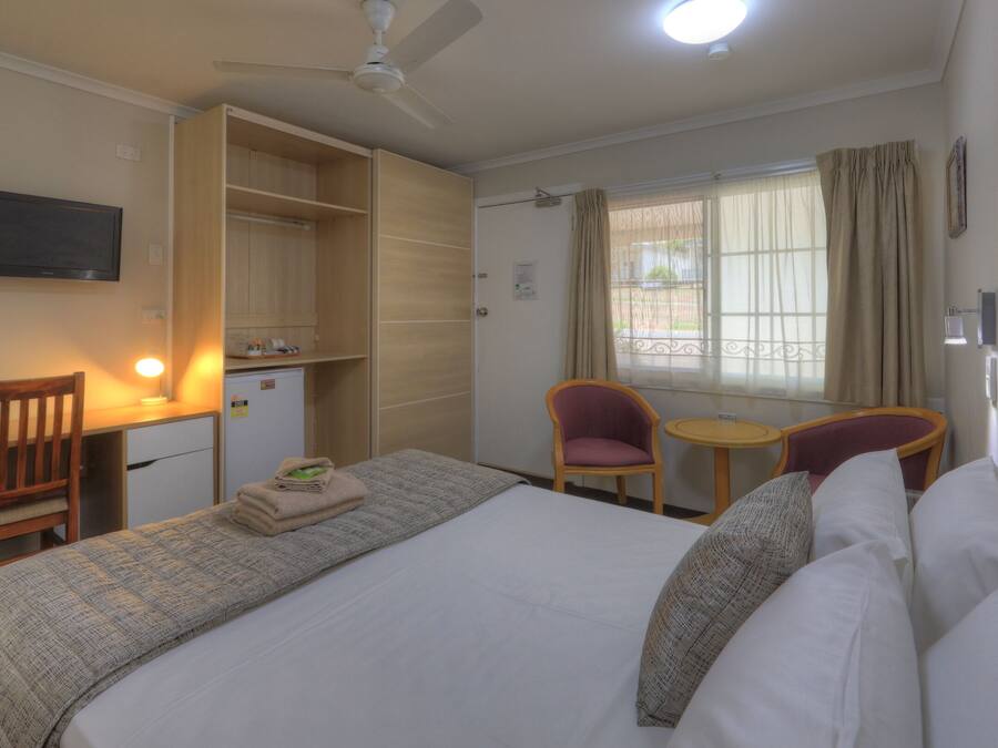 Quarto de hotel com cama, escrivaninha, cadeira, mesa pequena, ventilador de teto e janela com cortinas.
