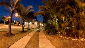 Caminho bem iluminado que leva a um edifício de pedra, com um jardim ajardinado e palmeiras.