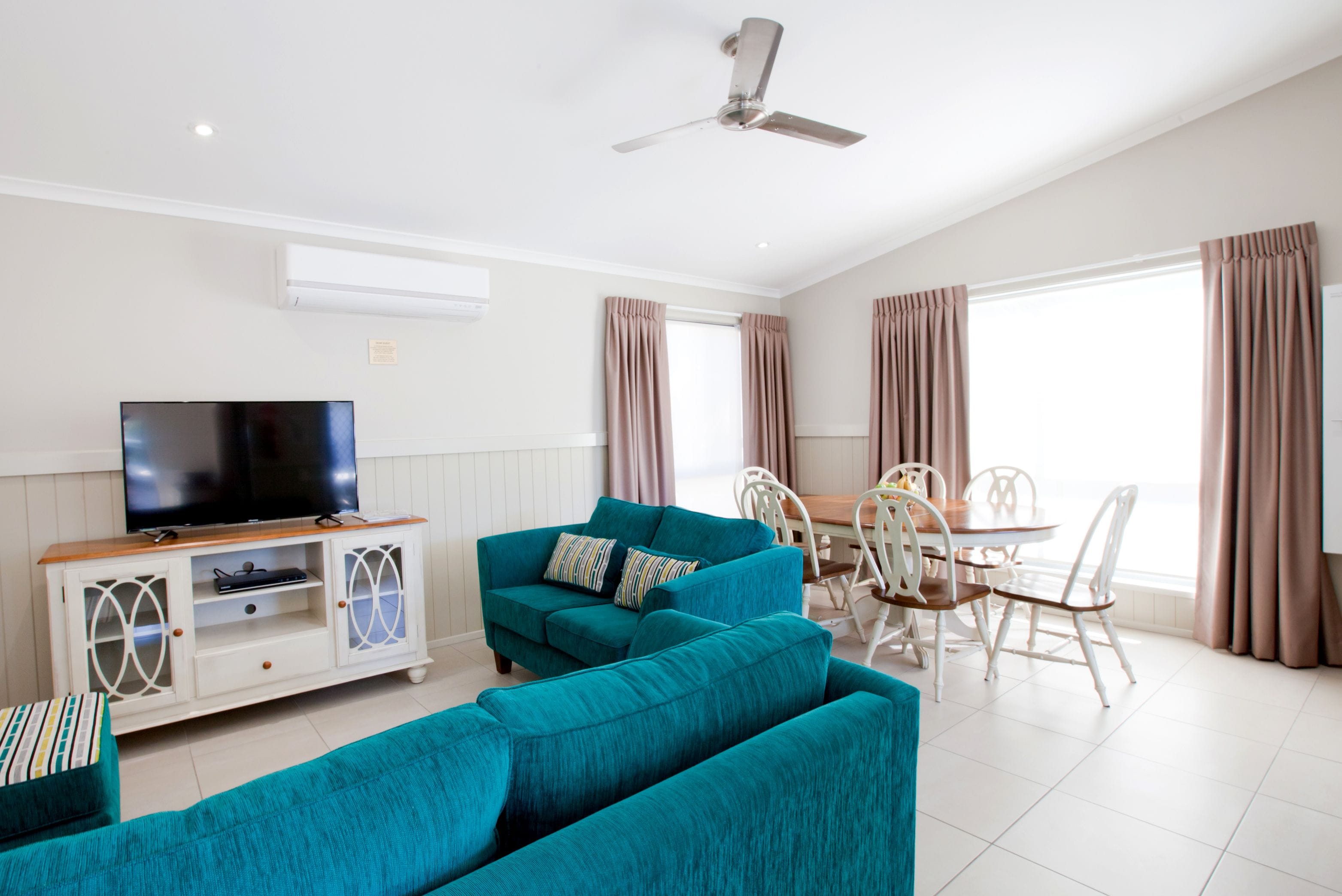 Uma sala de estar moderna com sofás azuis-esverdeados, uma TV sobre um armário branco, uma mesa de jantar com cadeiras e janelas amplas com cortinas.