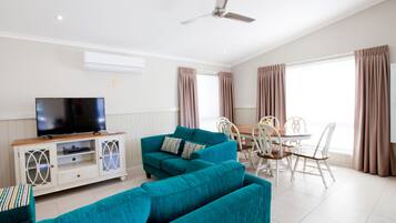 Uma sala de estar moderna com sofás azuis-esverdeados, uma TV sobre um armário branco, uma mesa de jantar com cadeiras e janelas amplas com cortinas.
