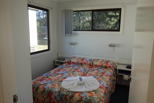 Quarto pequeno com uma cama de solteiro, edredom floral, duas mesas de cabeceira, janela com persianas e vista para árvores.