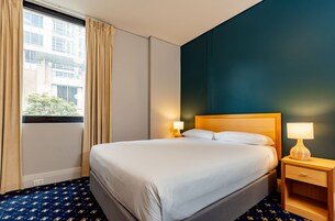 Quarto de hotel com uma cama, duas mesas de cabeceira, uma janela com cortinas e vista para edifícios.