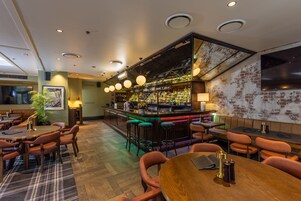 Uma área de bar com balcão de madeira, banquetas e diversos tipos de garrafas. Há mesas e cadeiras para sentar, e uma tela grande na parede.