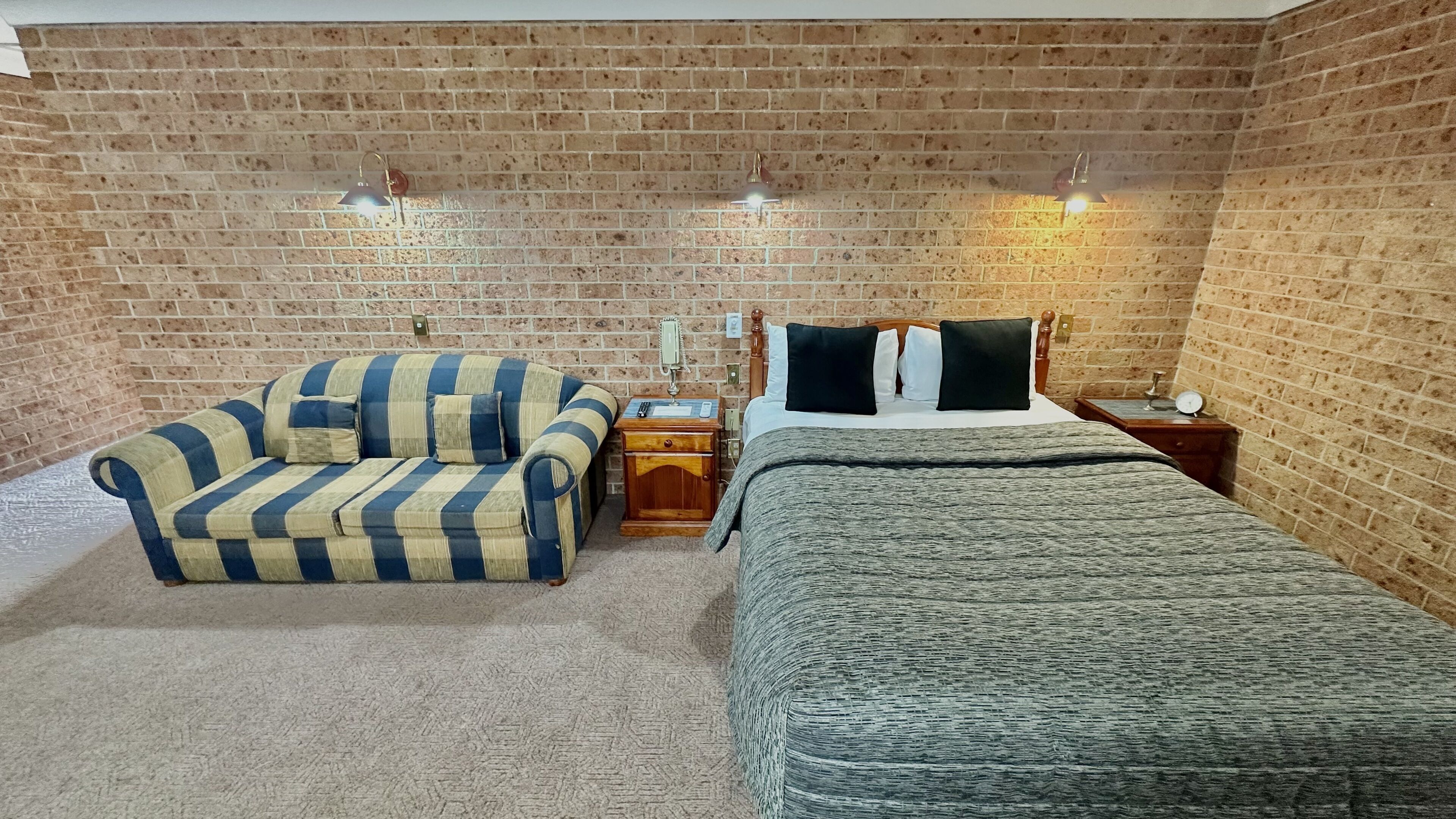 Quarto com parede de tijolos, um sofá listrado, uma cama com manta cinzenta e duas mesas de cabeceira com abajures.
