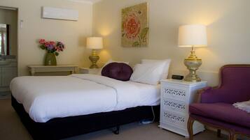 Quarto de hotel com uma cama, duas mesas de cabeceira com luminárias, uma poltrona roxa, um vaso com flores e um quadro na parede.