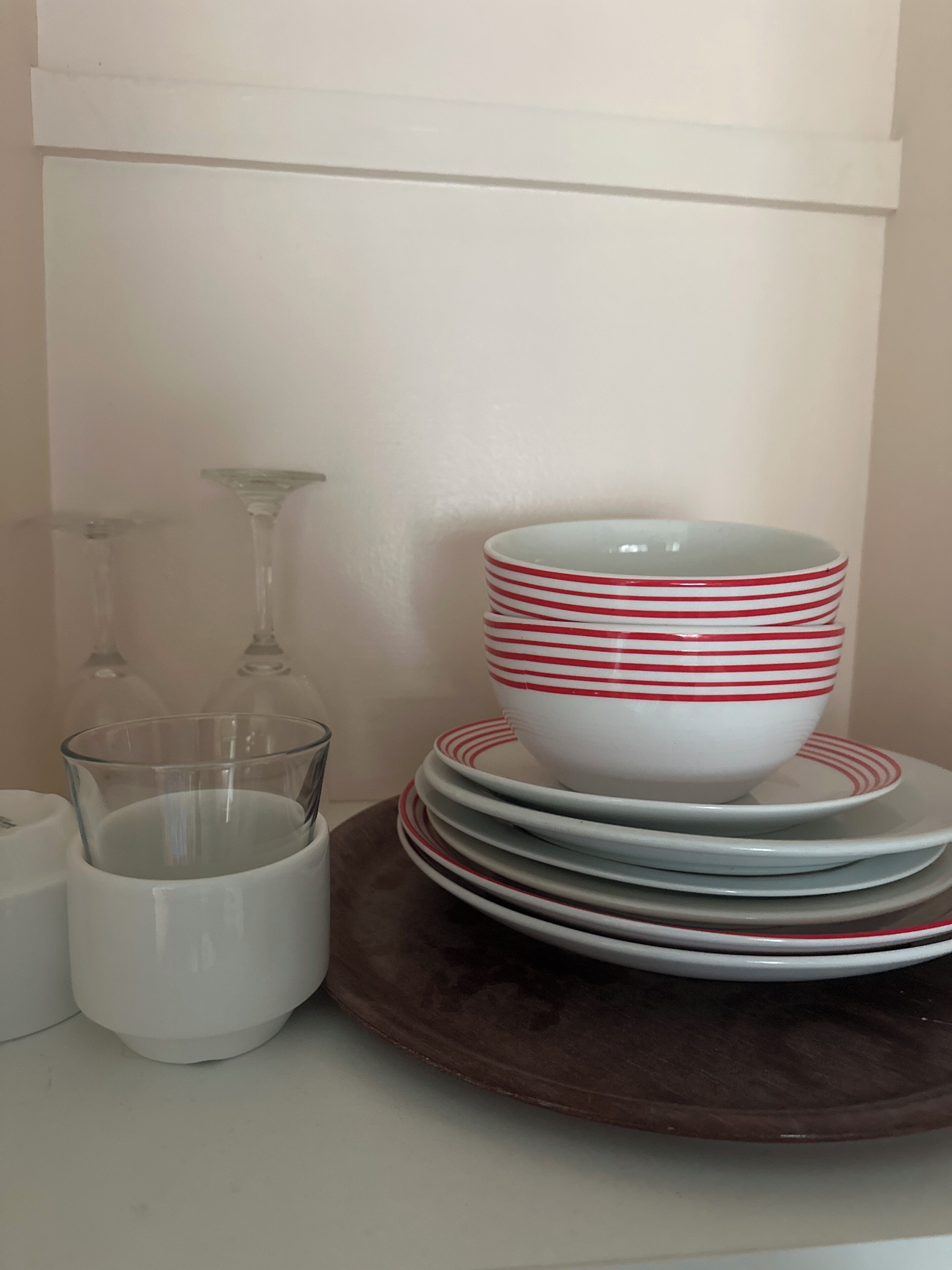 Uma pilha de tigelas e pratos listrados em branco e vermelho sobre uma bandeja marrom, com um jarro de vidro transparente e duas taças de vinho em uma superfície branca.