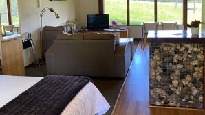 Quarto de hotel com cama, sofá, escrivaninha com computador e vista para um campo verdejante através de grandes janelas.