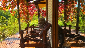 Um deck de madeira ao ar livre com pérgola, churrasqueira e mesa de vidro.
