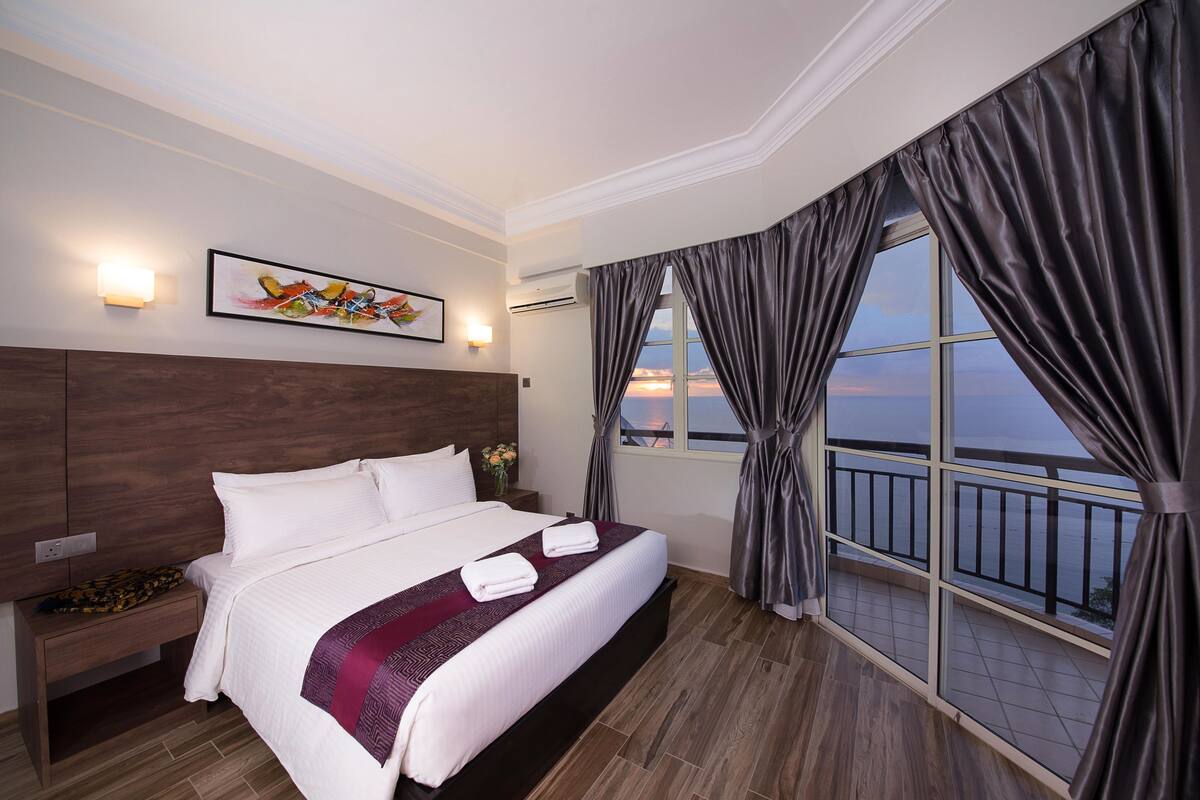 Quarto de hotel com uma cama grande, uma mesa de cabeceira, ar condicionado fixado na parede, uma obra de arte emoldurada e vista para o mar através de uma janela com cortinas.